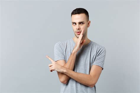 그의 손 뒤에 있는 우연한 옷 속 삭 임에서 젊은 남자의 초상화 옆에 회색 벽 배경에 고립 된 인덱스 손가락을 가리키는 사람들은 진심으로 감정 라이프 스타일 개념입니다 복사