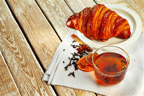 차가 있는 흰색 접시에 크루아상 오래된 나무 테이블에 차를 가진 머그잔 냅킨에 전체 잎 홍차 레몬과 계피 음식과 가까운 텍스트를 위한 공간과 여유 공간을 복사합니다 0명에