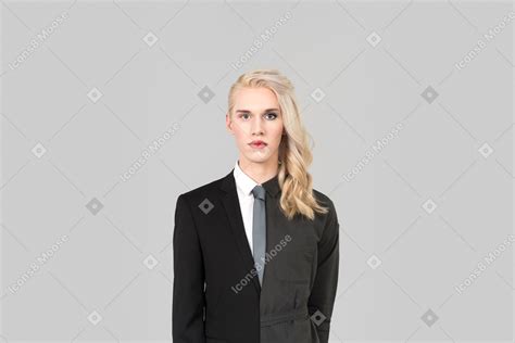 얼굴의 반은 화장으로 덮인 아름다운 트랜스젠더 반은 전통적인 여성의 모습 반은 남성의 옷을 입고 평범한 회색 배경에 서서 진지한 얼굴 표정을 기대하고 있습니다 사진