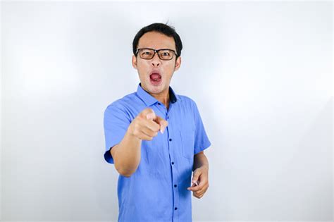 젊은 아시아 남자 입고 파란색 셔츠는 흰색 배경 위에 고립 된 카메라에서 소리지르고 손가락을 가리키는 재미있는 화가 얼굴입니다 20 29세에 대한 스톡 사진 및 기타 이미지
