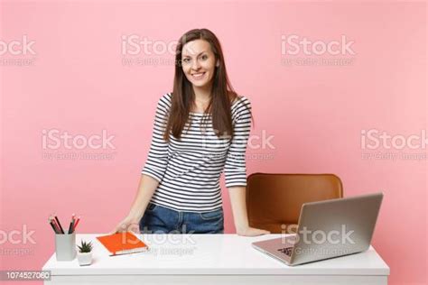 영 캐주얼 옷에서 여자 웃는 작동 파스텔 핑크 배경에 고립의 현대 Pc 노트북과 흰색 책상 근처에 서 성과 비즈니스 경력 개념입니다 광고에 대 한 공간을 복사 합니다 갈색