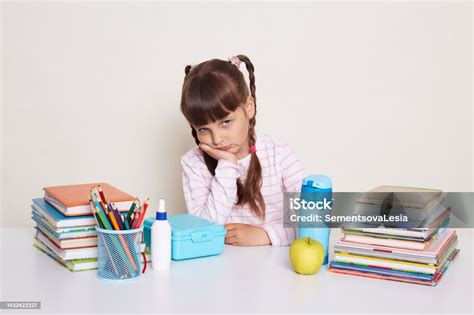 어두운 머리카락과 머리띠를 가진 슬픈 화가 난 어린 여학생이 책으로 둘러싸인 테이블에 앉아 턱 아래에 손을 대고 지루하고 지쳐서 카메라를 바라 보는 실내 장면 6 7 살에