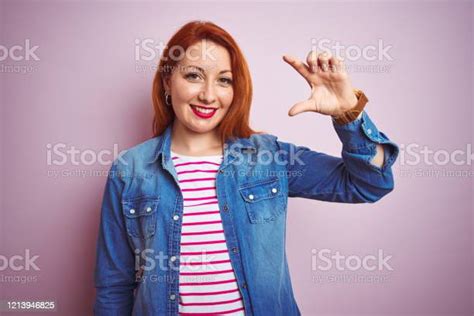 데님 셔츠와 스트라이프 티셔츠를 입은 아름다운 빨간 머리 여성은 손가락과 카메라를 보고 작은 크기의 기호를하고 손으로 미소와 자신감 몸짓 고립 된 분홍색 배경 위에 개념을