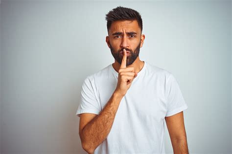 고립 된 흰색 배경 위에 서있는 티셔츠를 입은 젊은 인도 남자가 입술에 손가락으로 조용히 해달라고 요청했습니다 침묵과 비밀 개념 T 셔츠에 대한 스톡 사진 및 기타 이미지