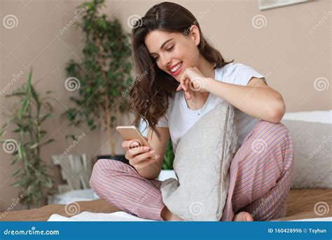 Linda Jovem Morena Sorridente Sentada Na Cama E Usando Um Telefone Em Seu Quarto Foto De Stock