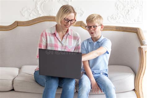 Счастливая молодая мама и ее сын сидя на софе и используя ноутбук Молодой мальчик показывая что