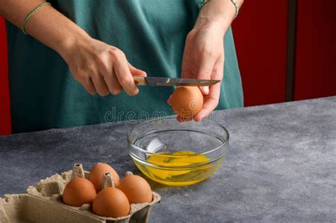 A Womanand X27s Hand Cracking An Egg With Knife Into A Clear Glass Bowl In The Gray Kitchen Table