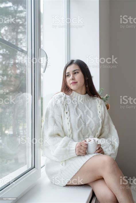 따뜻한 흰색 겨울 스웨터를 입은 여자는 크리스마스 이브에 집에서 창문 옆에 서서 마시멜로가 든 컵을 들고 전나무를 들고 있다