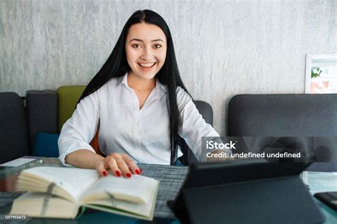 캐주얼 한 흰색 셔츠를 입은 젊은 예쁜 아시아 여성의 초상화 쇼핑 공간이나 카페에 앉아 태블릿과 책으로 작업하는 동안 카메라를 미소 짓고 보고 있습니다 갈색 머리에 대한