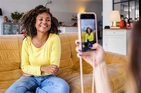 Woman Taking Photo Of Smiling Friend Sitting On Sofa At Home Stock