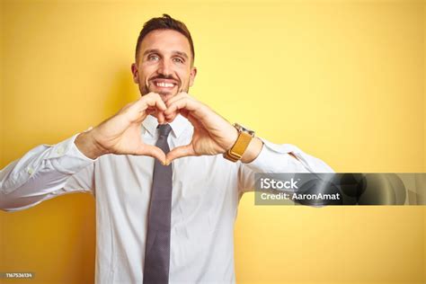 노란색 고립 된 배경 위에 우아한 흰색 셔츠를 입고 젊은 잘 생긴 사업가는 손으로 심장 기호 모양을하고 사랑에 미소 로맨틱 한