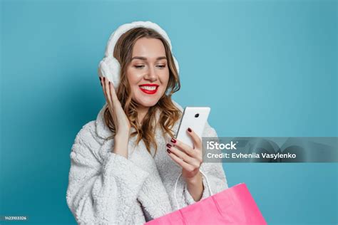 인공 흰 모피 코트와 모피 헤드폰을 입은 행복한 젊은 여성이 휴대 전화를 들고 메시지를 읽거나 블루 스튜디오 배경에 고립 된