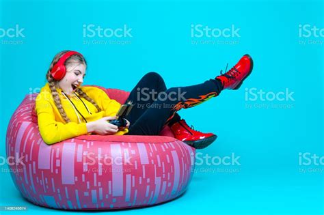 청소년 라이프 스타일 개념 청록색 배경에 놀란 표정으로 쿠션 의자에서 게임을 위해 핸드폰을 사용하는 동안 게임 패드 조이스틱과 후드 재킷에 Winsome 십대 소녀 14