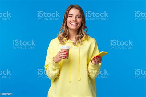 카페 근처에 서있는 쾌활한 젊은 여학생은 테이크 아웃 커피를 주문하고 종이 컵을 마시고 휴대 전화를 들고 하얀 미소를 지으며 카메라를 보고 만족합니다 교육에 대한 스톡 사진