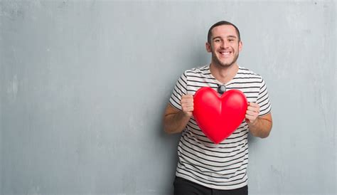 서 치아를 보여주는 자신감 미소로 웃 고 행복 한 얼굴로 붉은 마음을 잡고 회색 그런 지 벽에 젊은 백인 남자 건강한 생활방식에 대한 스톡 사진 및 기타 이미지 Istock