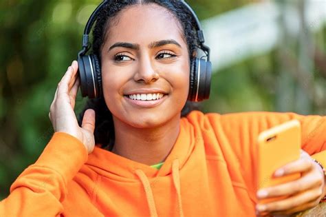 무선 헤드폰과 휴대전화로 음악을 듣고 있는 오렌지색 후드티를 입은 아름다운 혼혈 아프리카계 미국인 소녀 십대 여성 젊은 여성의 야외 초상화 사진 배경 및 무료 다운로드를위한