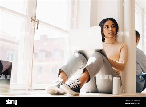 Full Length Of Female Computer Programmer Looking Away While Sitting By Window In Office Stock