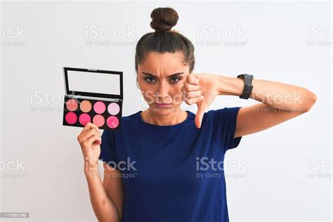 화가 얼굴 엄지 손가락으로 싫어하는 것을 보여주는 부정적인 기호 거부 개념으로 고립 된 흰색 배경 위에 서있는 메이크업을 들고있는 젊은 아름다운 여성 갈색 머리에 대한 스톡