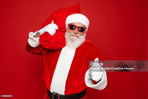 빨간 배경에 고립 된 카메라를 보면서 제스처와 같은 엄지 손가락을 위로 손을 보여주는 자루 가방과 의상을 입고 행복 흥분 재미 있은 수염을 기른 늙은 산타 클로스 12월에