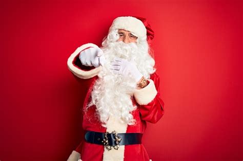 고립된 빨간 배경 위에 서있는 산타 의상을 입은 중년 잘생긴 남자가 당신을 비웃고 손가락을 입에 대고 카메라를 가리키며 부끄러운 표정을 짓고 있습니다 12월에 대한 스톡