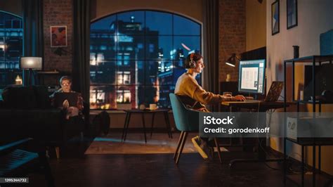 젊은 부부는 집에서 시간을 보내고 저녁에는 세련된 로프트 아파트에서 컴퓨터로 일합니다 소파에서 쉬고 노트북에서 소셜 미디어를 탐색하는 여성 Ux 인터페이스를 디자인하는 사람