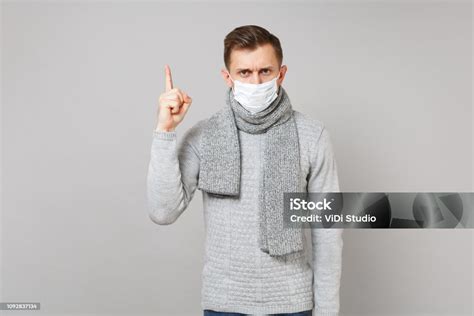 회색 스웨터에 젊은 남자 스카프 메 마른 얼굴 마스크 검지 손가락을 가리키는 회색 벽 배경에 고립 건강 한 생활 습관 병 아픈 질병 치료 추운 계절 개념 복사 공간을 비웃는