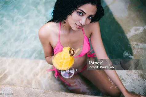 다과를 위해 신선한 음료를 들고 수영장에서 분홍색 수영복을 입은 갈색 머리 여성의 위 사진에서 휴가 기간 동안 빌라 리조트에서 휴식을 즐기는 슬림 한 인물을 가진 매력적인