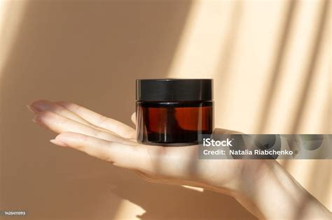 여성의 손바닥에는 갈색 배경에 호박색 유리로 만든 보습 크림 항아리가 있습니다 노화 방지 마스크 화장품 스크럽으로 라벨링하지 않고 병을 모형합니다 스킨케어 컨셉 쥠에 대한