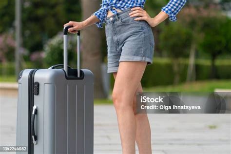 반바지와 엉덩이에 손을 얹은 셔츠를 입은 젊은 날씬한 여성은 복사 공간이 있는 회색 가방을 클로즈업하여 옆으로 서 있다 여행 개념 가냘픈에 대한 스톡 사진 및 기타 이미지