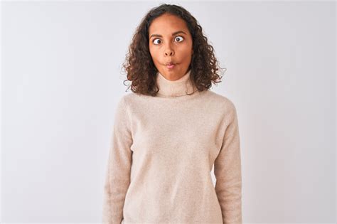 고립 된 흰색 배경 위에 서있는 터틀넥 스웨터를 입은 젊은 브라질 여성은 입술 미친 코믹 한 제스처로 물고기 얼굴을 만듭니다