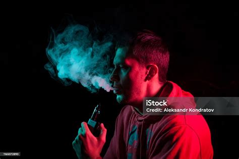 성인 심각한 남자의 얼굴의 초상화를 닫아 녹색 독성 연기를 내뿜고 전자 담배를 피우고 검은 배경에 파란색과 빨간색 으로 빛나는 Vape 건강에 해를 끼칩니다 검은색에 대한