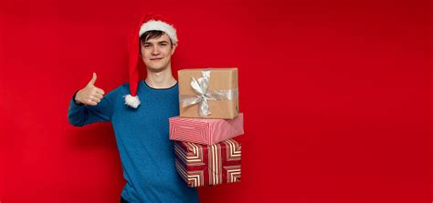 산타 모자를 쓴 청년이 크리스마스 선물을 들고 다른 한 손은 엄지 손가락을 위로 향하게합니다 감정에 대한 스톡 사진 및 기타 이미지 Istock