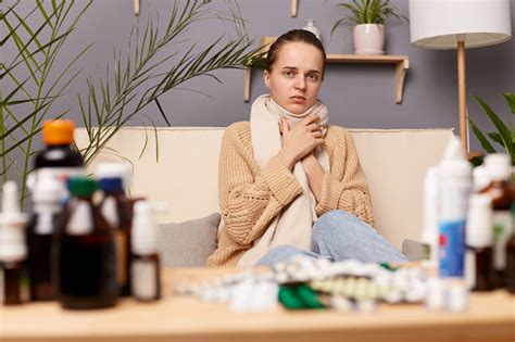 따뜻한 점퍼를 입고 스카프에 싸인 아픈 건강에 해로운 여성의 실내 촬영 기침에 앉아 목을 만지고 목이 아프고 끔찍한 고통을