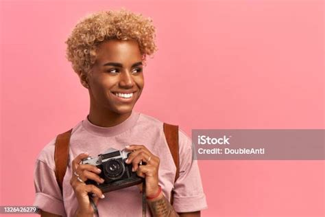 재능있는 아프로 여성 사진 작가의 이미지는 모험 여행 중에 전문 적인 사진을 찍고 복고풍 카메라를 사용하고 라벤더 티셔츠와 배낭을 입고 휴일을 즐기고 실내 포즈를 취합니다