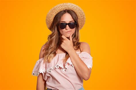 사려 깊고 생각에 잠겨있는 아름 다운 여성 턱을 보유 하 고 주저 식 그늘 및 여름 모자 어깨 소매 블라우스를 입은 오렌지 배경에 포즈를 착용 사람과 나머지 개념 계절에