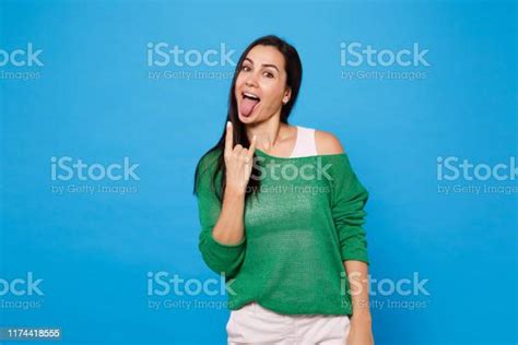 스튜디오의 밝은 파란색 벽 배경에 고립 된 중금속 바위 표지판을 묘사 한 혀를 보여주는 녹색 캐주얼 옷을 입은 쾌활한 젊은 여성의 초상화 사람 라이프 스타일 개념입니다 복사