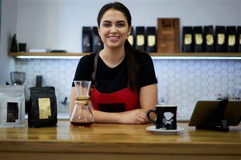 매력적인 긍정적인 젊은 여자의 반 길이 초상화에서 서 있는 빨간 앞치마를 입은 바와 카메라를 보고 세련 된 커피숍 인테리어에서