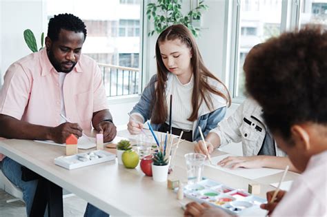 젊은 아프리카 교사 나 교사는 의자에 앉아 백인 학생이나 학교에서 교실에서 학생들을위한 예술의 수채화와 그림과 그림을 가르치고있다 아티스트 컨셉교육 가족에 대한 스톡 사진
