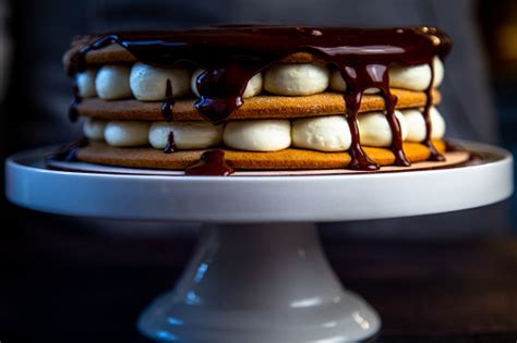 갈색 꿀 케이크 화이트 크림을 곁들인 케이크 옆구리를 흐르는 다크 초콜릿을 얹은 케이크가 어두운 배경의 흰색 스탠드위에 서 있습니다 갈색에 대한 스톡 사진 및 기타 이미지