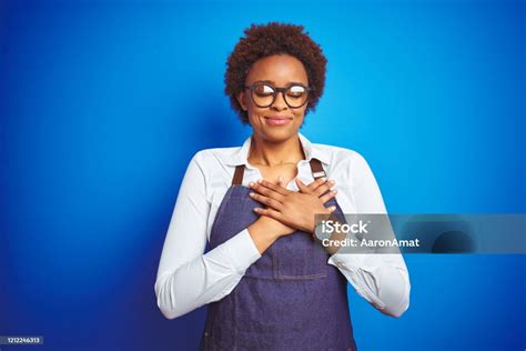 파란 배경 위에 비즈니스 앞치마를 입은 젊은 아프리카 계 미국인 여성 상점 주인은 닫힌 눈과 얼굴에 감사하는 제스처로 가슴에