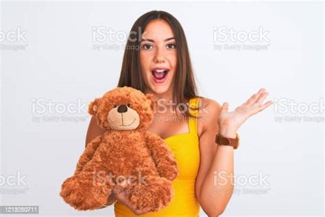 고립 된 흰색 배경 위에 서있는 귀여운 테디 베어를 들고있는 젊은 아름다운 소녀는 매우 행복하고 흥분 큰 미소와 함께 비명을