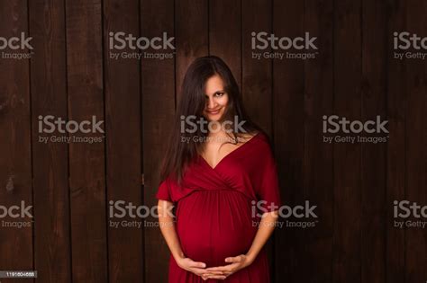 빨간 드레스를 입은 임산부는 어두운 갈색 배경에 배에 손을 들고 있습니다 임신 출산 기대 개념 임신의 아름다운 부드러운 분위기 사진 갈색 머리에 대한 스톡 사진 및 기타