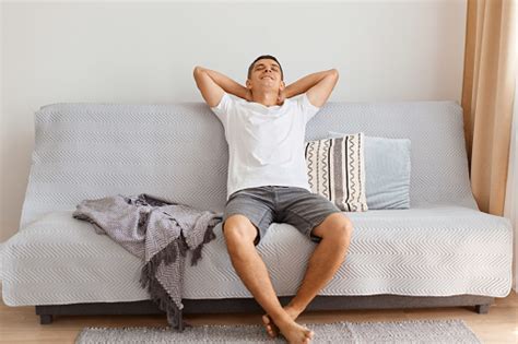 거실에서 기침에 앉아 하얀 티셔츠를 입은 편안한 만족한 남자의 초상화가 손을 들고 긍정적인 미소를 지으며 눈을 감고 즐거운 순간을 기억합니다 35 39세에 대한 스톡 사진