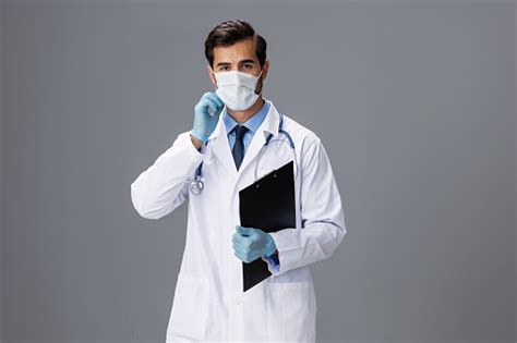 흰 가운을 입고 의료용 마스크를 쓴 남자 의사가 손에 메모장을 들고 회색의 고립된 배경 복사 공간 텍스트를 위한 공간에서 카메라를 바라본다 25 29세에 대한 스톡 사진 및