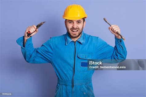 노란 헬멧과 파란색 유니폼을 입고 화가 난 건설 노동자 엔지니어의 초상화를 닫고 손을 들고 분노로 무언가를 외치고 수염이 있는 남자가 두 손으로 렌치 도구를 들고 있다 건설