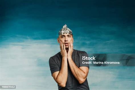 호일 모자를 쓴 한 남자가 파란 배경에 눈과 입을 벌린 채 두 손을 무섭게 잡는다 모자 모자류에 대한 스톡 사진 및 기타 이미지