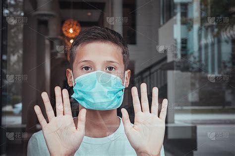 在covid 19冠状病毒全球大流行期间，患病的小男孩戴着防护口罩在家，躲在隔离和封锁的窗户后面，看