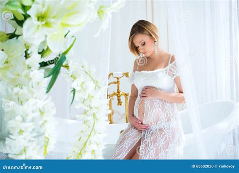 Беременная девушка в белом платье шнурка сидя в интерьере с цветками и объятия Belly Стоковое