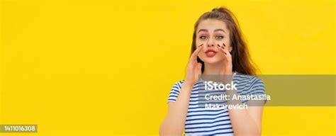 친근한 쾌활한 갈색 머리 소녀가 큰 소리로 외치고 메가폰에서 손을 결합하고 노란색 배경 위에 흰색 파란색 줄무늬 티셔츠에 서서