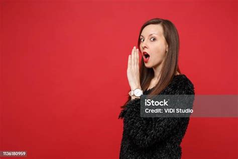 밝은 빨간색 벽 배경 스튜디오 초상화에 고립 된 손 제스처와 검은 모피 스웨터 속 삭 임 비밀에 놀란 젊은 여자 사람들은